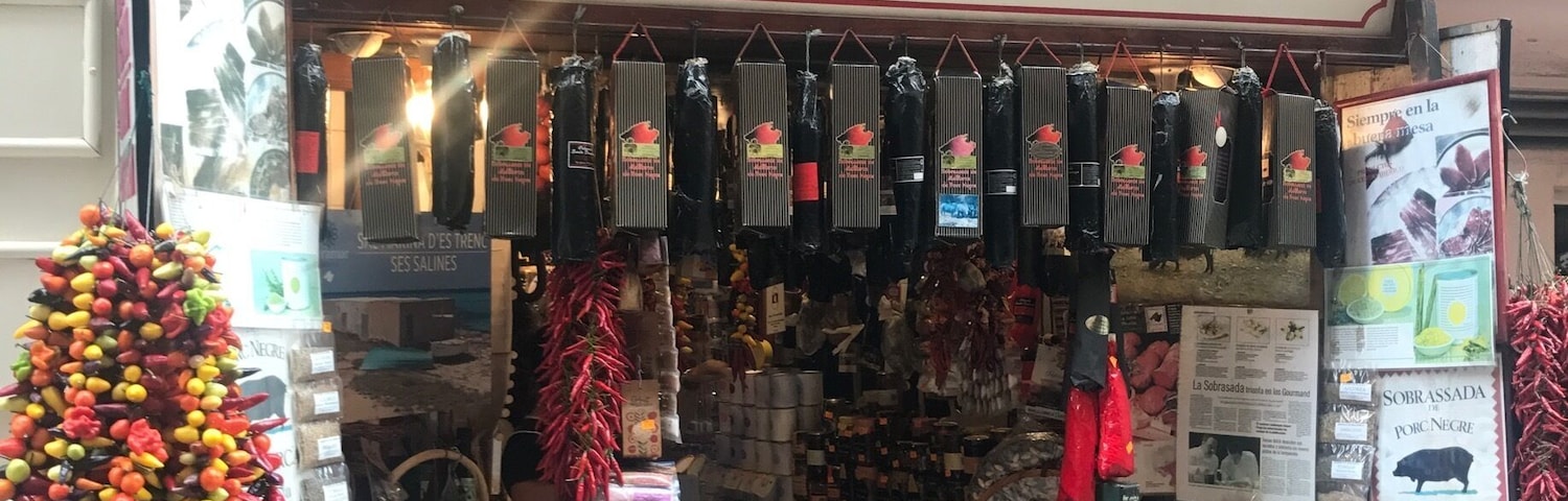 Uma banca de mercado chamada 'SOBRASADA - COLUMADO STO. DOMINGO' com diversos produtos expostos, como enchidos, frutas e legumes.