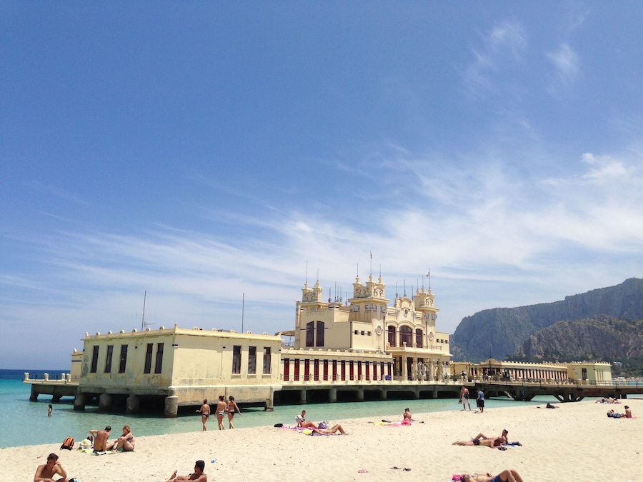 Uma praia com pessoas se banhando e tomando sol, um edifício amarelo grande com torres e uma montanha rochosa ao fundo.