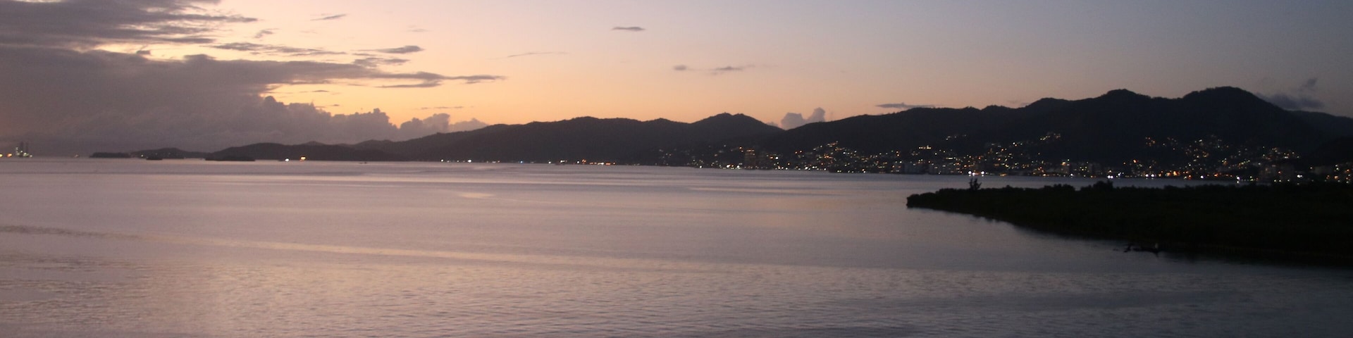 Água calma com um horizonte urbano ao longe, montanhas ao fundo e céu claro.