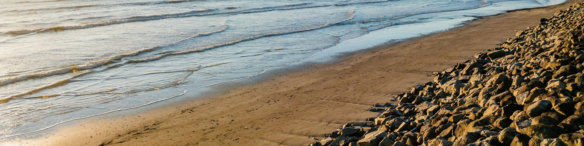 Uma praia rochosa com ondas, céu claro e montanhas ao longe.