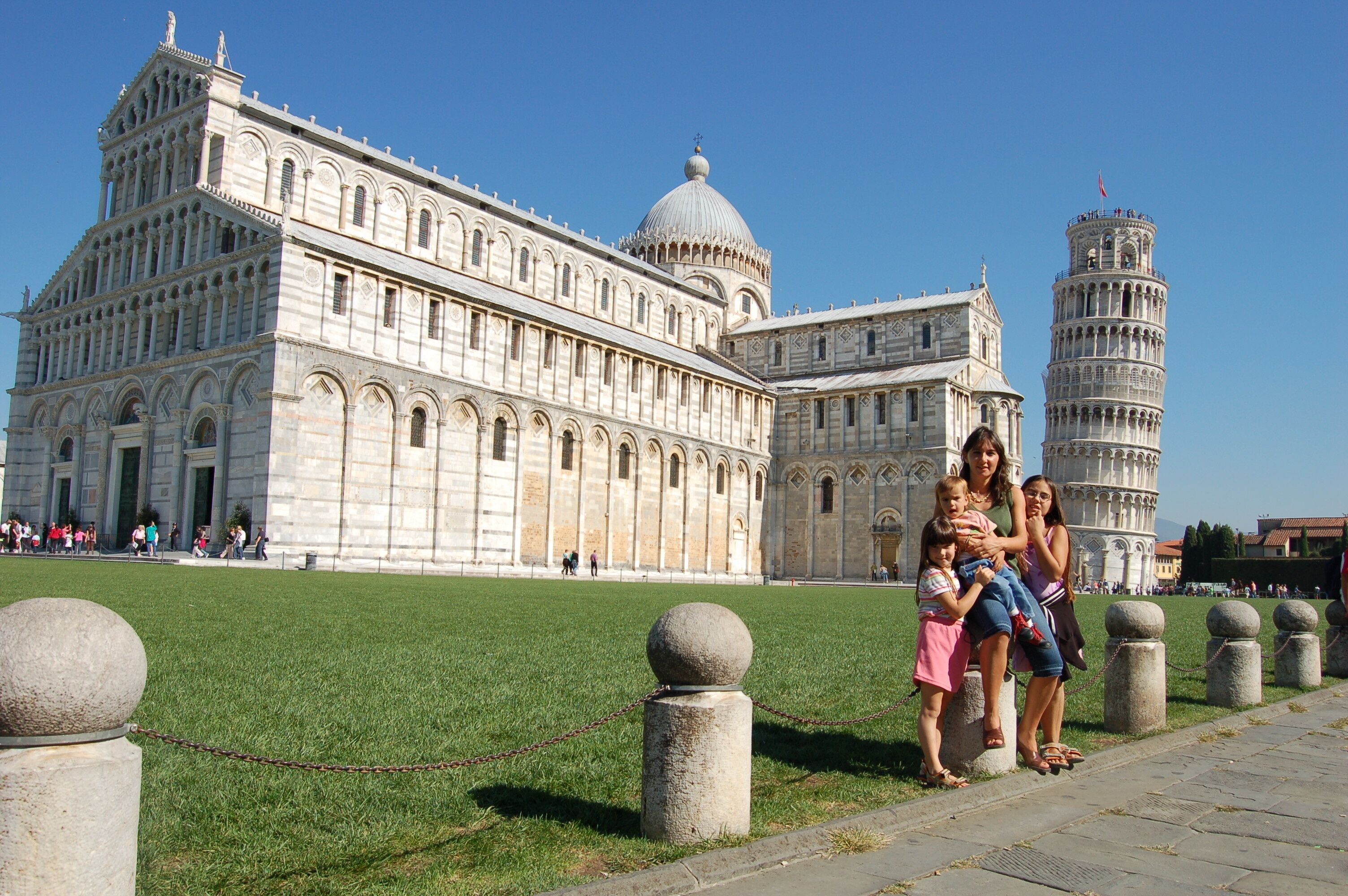 A Torre de Pisa inclinada, a Catedral de Pisa e uma área gramada com turistas.