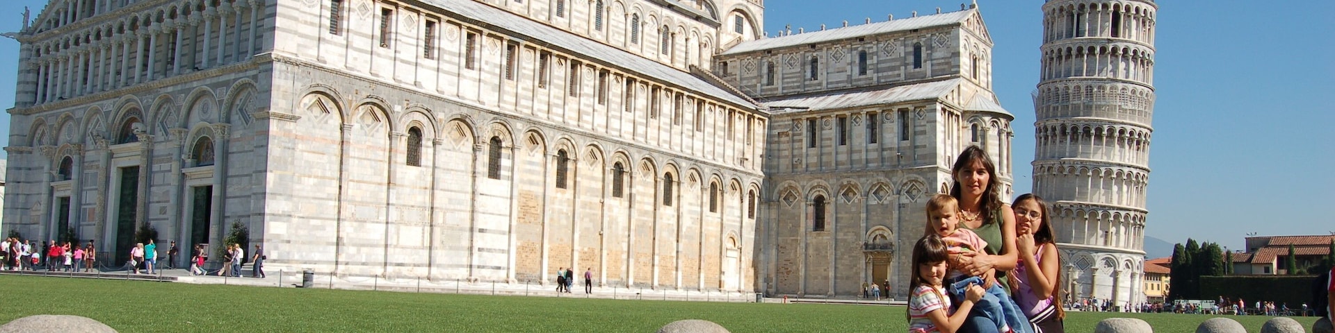 A Torre de Pisa inclinada, a Catedral de Pisa e uma área gramada com turistas.