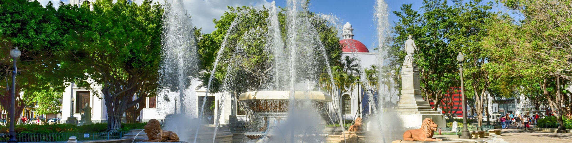 Uma fonte com vários jatos de água, rodeada por árvores e um edifício com cúpula vermelha.
