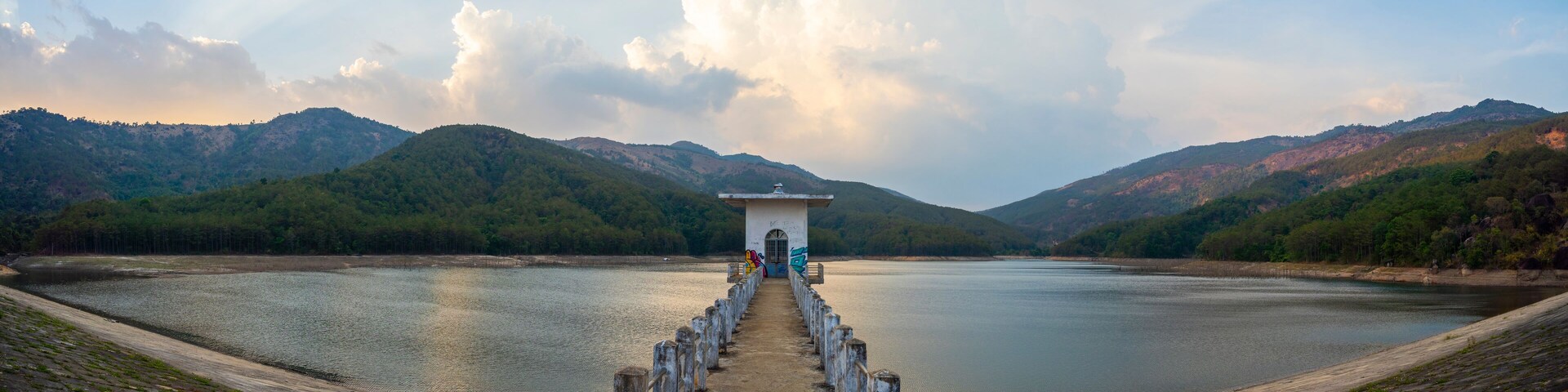 Uma barragem com uma plataforma de observação que dá para um rio calmo, rodeado por montanhas.