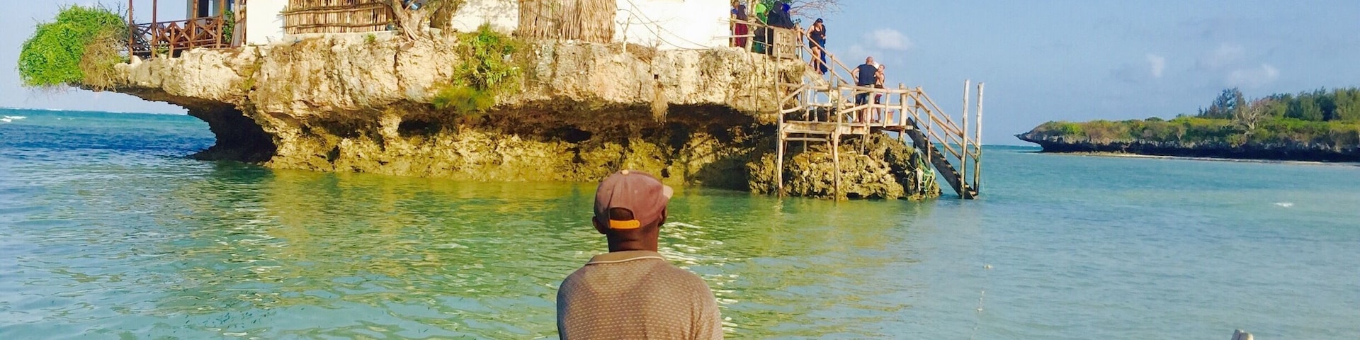 Um homem em um barco se aproximando de uma casa com telhado de palha em um afloramento rochoso à beira-mar.