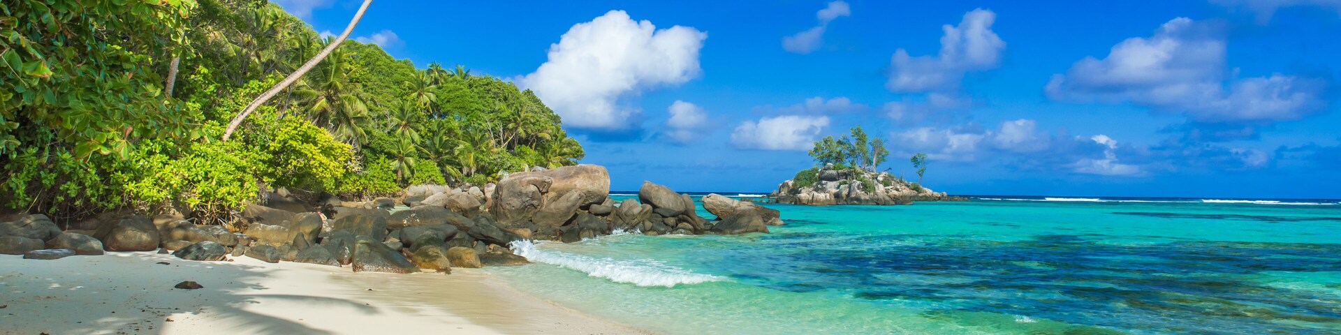 Uma praia tropical com águas turquesa cristalinas, areia branca, palmeiras e um afloramento rochoso ao longe.