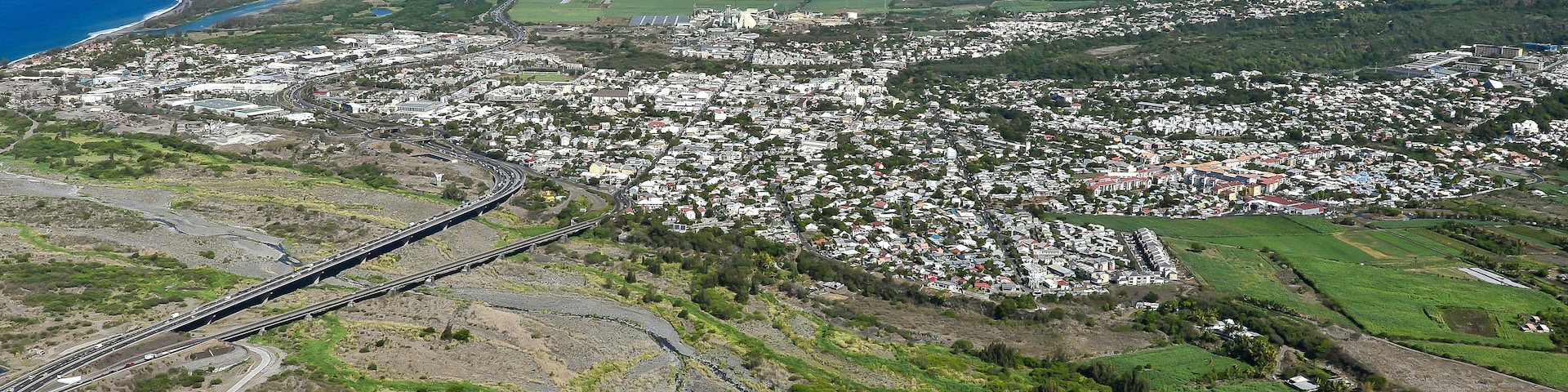 Uma vila costeira com uma ponte ligando ao continente, rodeada por colinas verdes e um céu azul límpido.