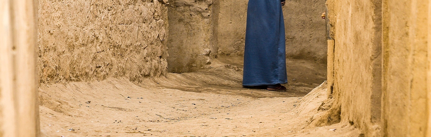 Um beco estreito com uma pessoa vestindo manto azul e lenço de cabeça, caminhando para longe da câmera.