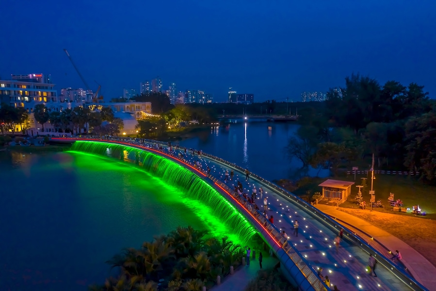 Vista noturna de uma paisagem urbana com uma ponte iluminada sobre um rio, rodeada por edifícios e palmeiras.