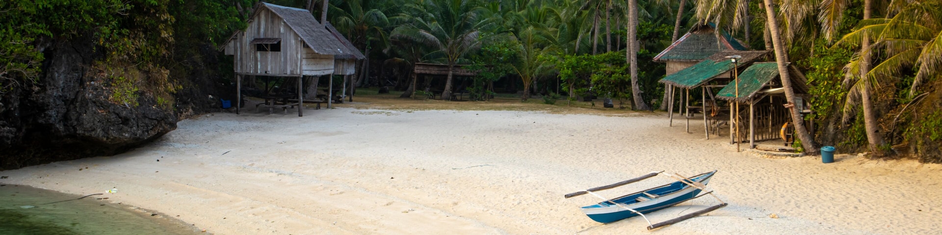 Uma praia com uma cabana de madeira, palmeiras e um barco na areia.