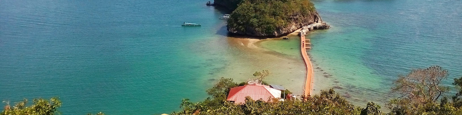 Paisagem costeira com várias ilhas pequenas cercadas por águas azul-turquesa. Um edifício com telhado vermelho é visível na margem, e um barco branco está ancorado perto.