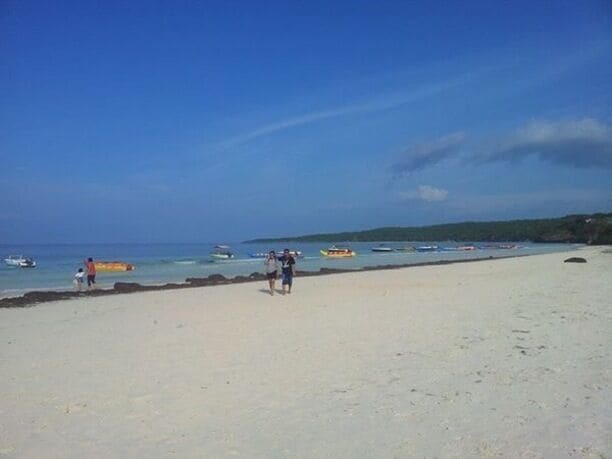 Uma praia arenosa com pessoas caminhando, barcos na água e céu claro.