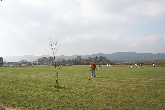 Um campo gramado com pessoas praticando um esporte, uma pequena árvore e colinas ao longe.