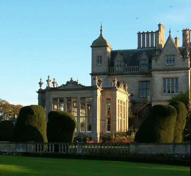 Um edifício grande e histórico com fachada simétrica, várias chaminés e um jardim bem cuidado, com sebes aparadas.
