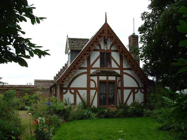 Uma casa em estilo Tudor com telhado íngreme, paredes brancas com vigas de madeira escura e um jardim bem cuidado com diversas plantas e flores.