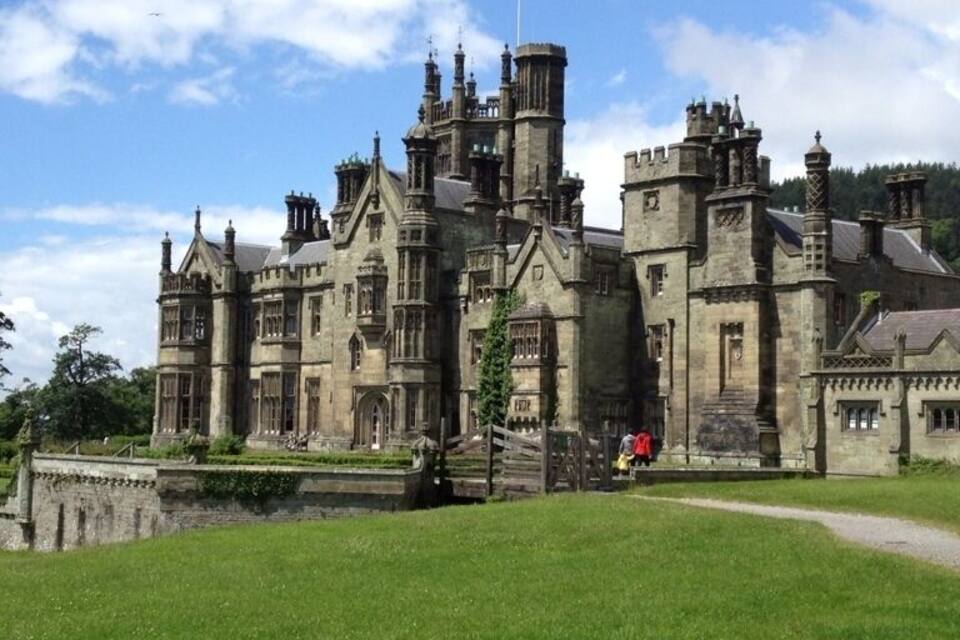 Um edifício grande e histórico de pedra, com várias torres e ameias, rodeado por um jardim bem cuidado e um muro de pedra.