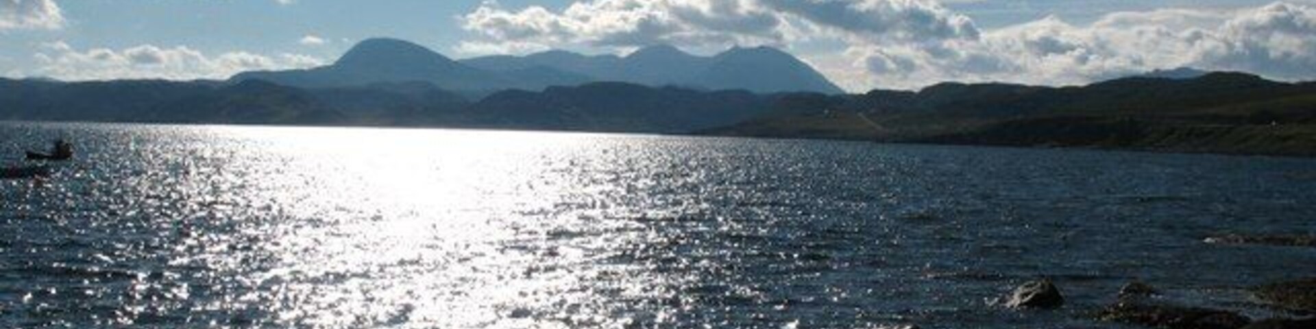 Um lago calmo com o sol refletindo na água, rodeado por montanhas e margens rochosas.