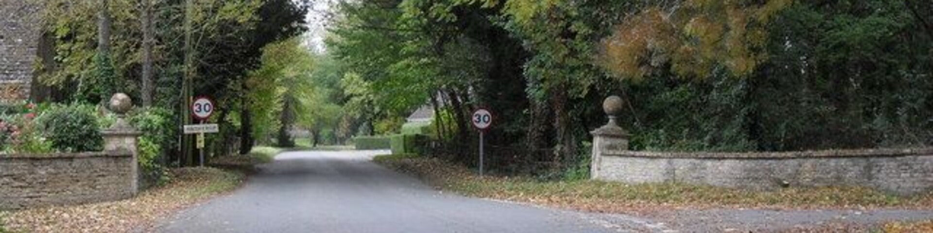 Uma estrada rural tranquila ladeada por árvores e muros de pedra, com uma placa de velocidade indicando 30 milhas por hora.