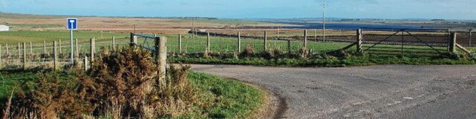 Uma estrada rural serpenteando para a direita, ladeada por campos abertos e uma cerca distante.