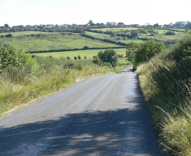 Uma estrada rural estreita, ladeada por campos gramados e árvores dispersas.