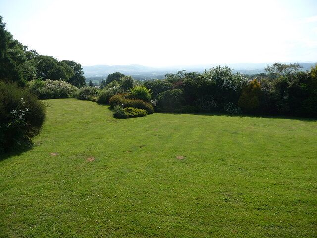 Um campo gramado com arbustos e árvores, céu claro e montanhas ao longe.