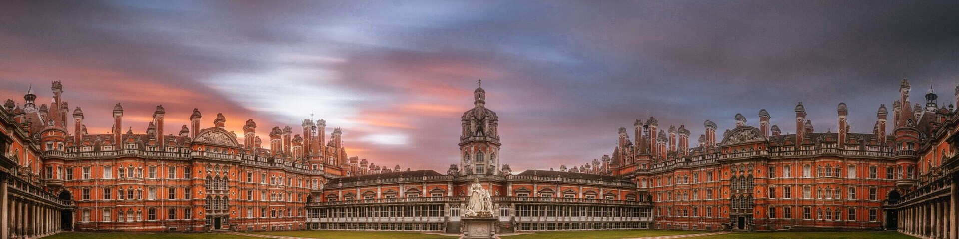 Um edifício imponente de tijolos vermelhos, com uma torre central e alas simétricas, rodeado por um gramado bem cuidado.