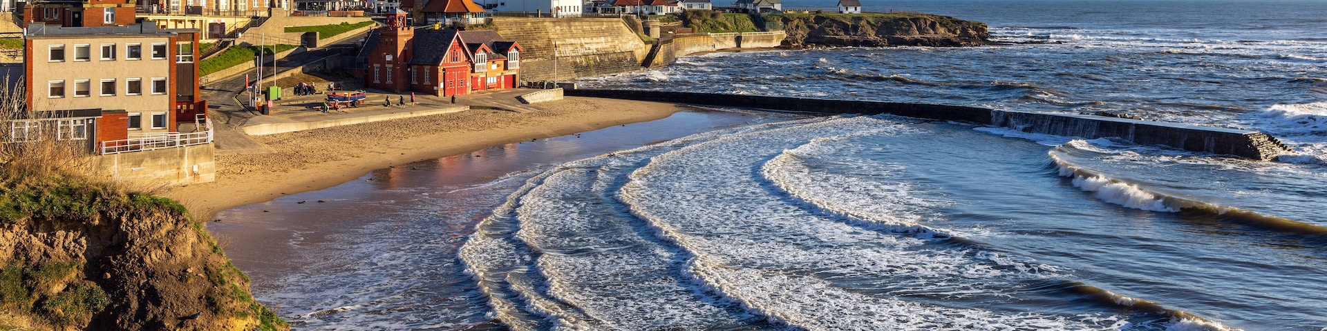 Uma vila costeira com praia de areia, um muro de contenção e edifícios à beira-mar.