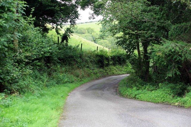 Uma estrada rural estreita e sinuosa, rodeada por vegetação exuberante e árvores.