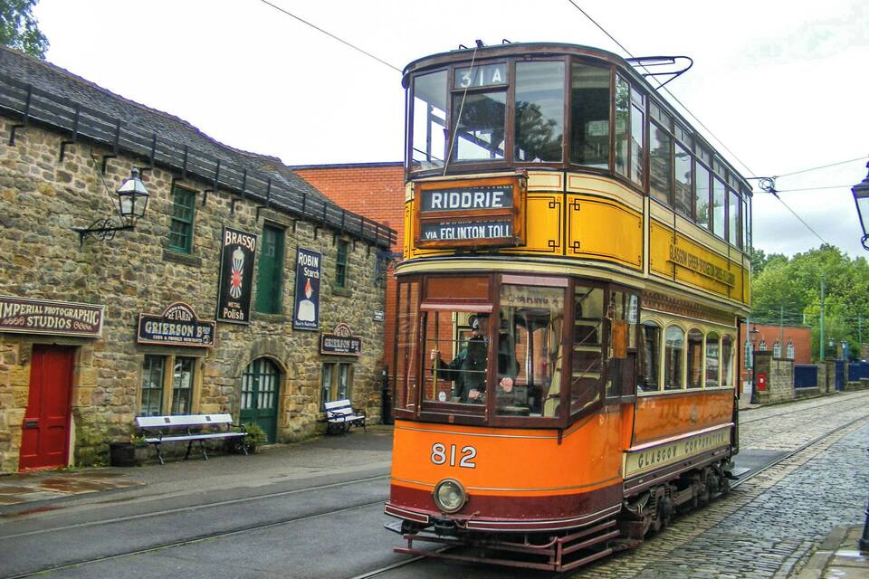 Um elétrico de dois andares laranja antigo, com o número 812, com a inscrição 'Riddrie' e 'Via Fglinton Toll', percorre uma rua de paralelepípedos, passando por edifícios históricos com placas de 'Brasso', 'Robin Starch' e 'Imperial Photographic Studios'.