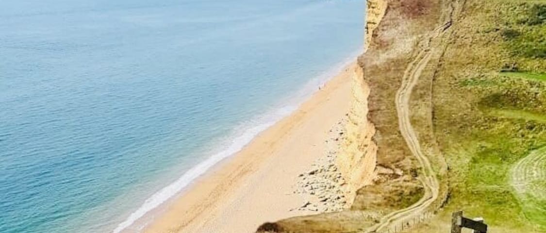 Paisagem costeira com uma praia de areia, um penhasco e um caminho de terra que leva a um portão de madeira.