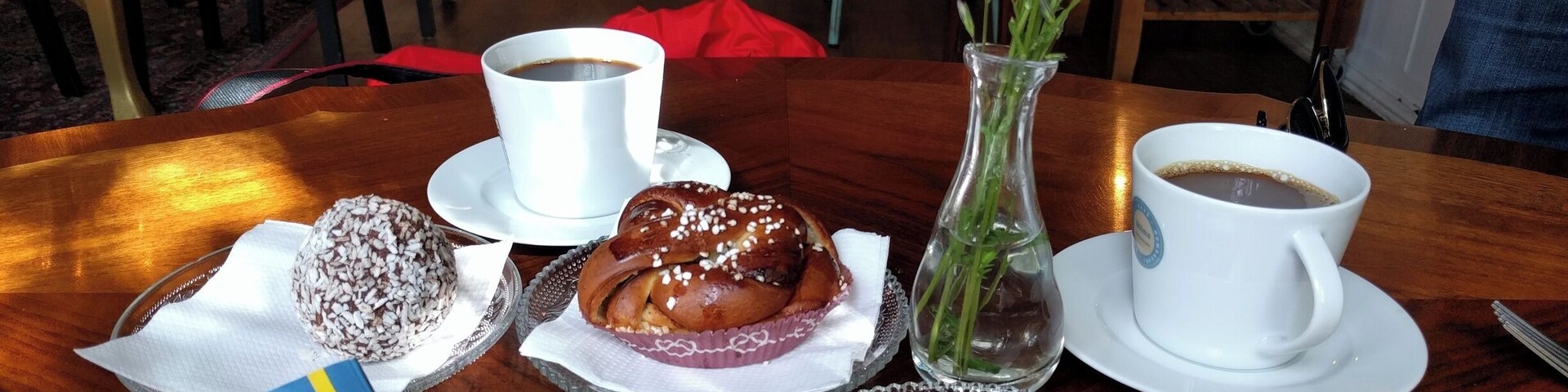Uma mesa com um pastel, um bolo com morangos, um copo de café e um vaso com flores.
