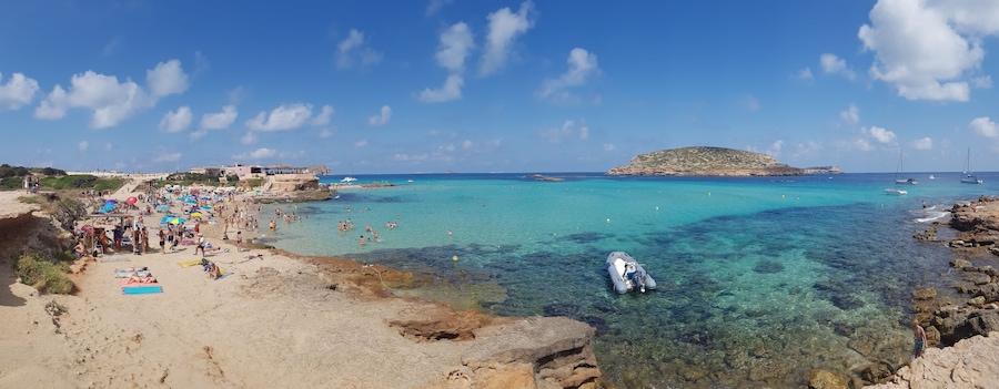 Uma praia lotada com águas turquesa cristalinas, costas rochosas e uma pequena ilha ao longe.