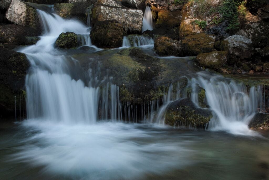 Uma cascata desce sobre rochas cobertas de musgo em uma área arborizada.