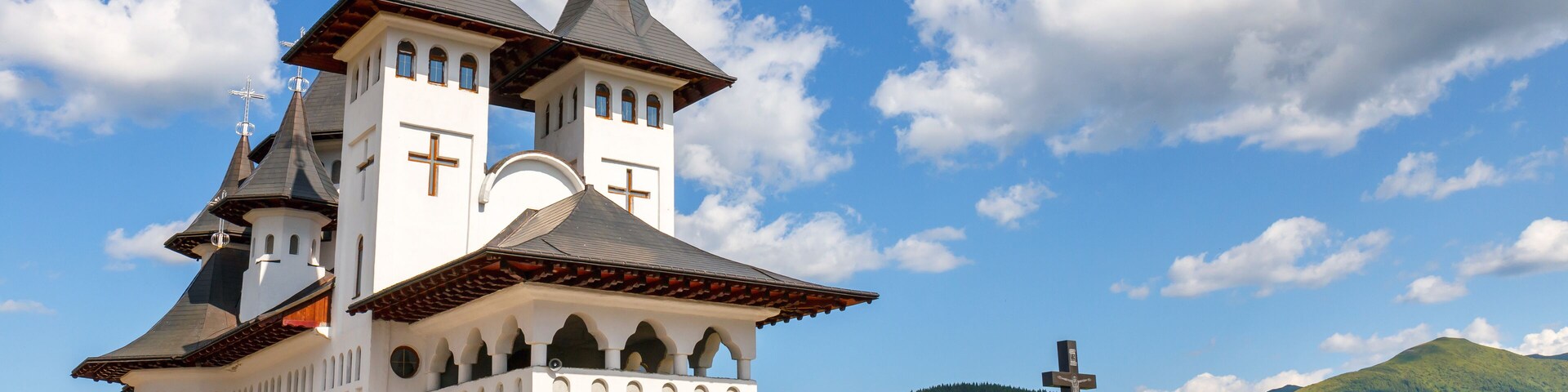Uma igreja branca com múltiplices torres e uma cruz no topo, emoldurada por uma paisagem montanhosa.