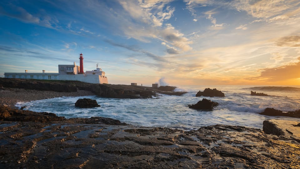 Farol, linha costeira rochosa e ondas quebrando nas rochas ao pôr do sol.