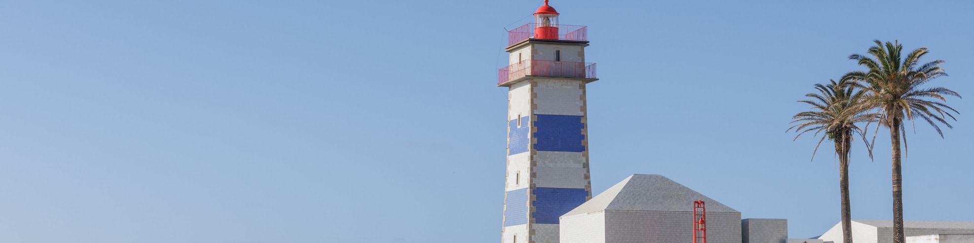 Um farol com a parte superior vermelha, um edifício branco com uma faixa azul e palmeiras à beira-mar.