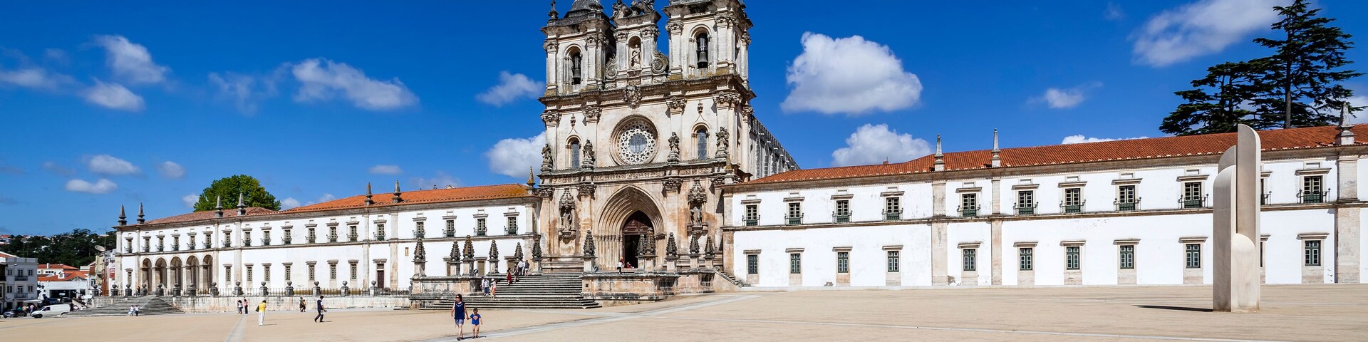 Um edifício histórico com uma torre imponente, uma grande praça aberta e um céu azul claro.