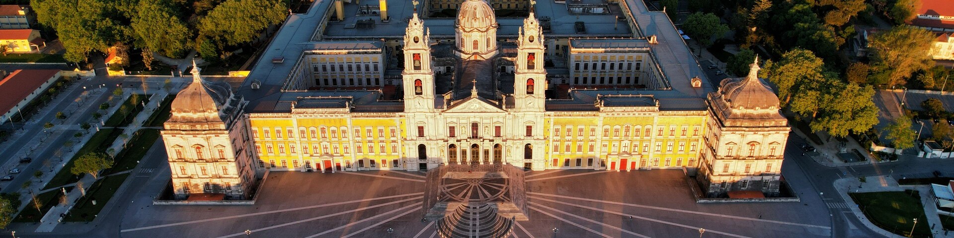 Vista aérea de um edifício imponente com duas cúpulas, cercado por uma ampla praça e uma área florestal densa.