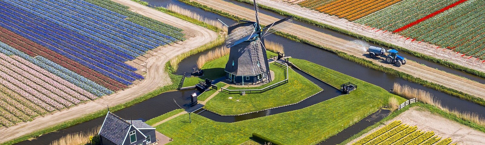 Vista aérea de um moinho de vento cercado por campos agrícolas, um pequeno edifício e um rio.