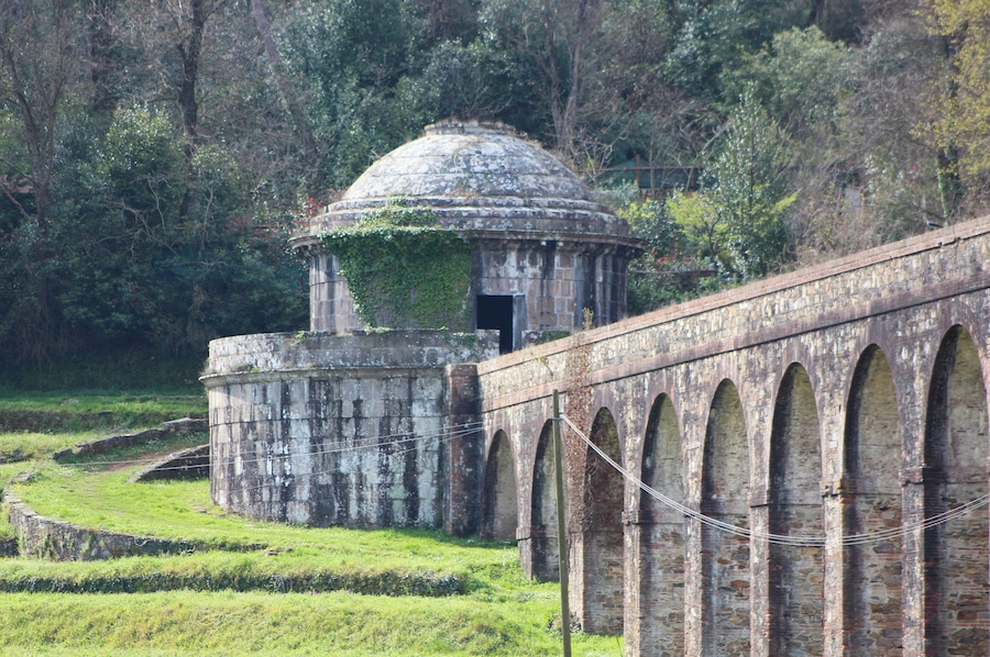 Uma ponte de pedra com arcos e uma estrutura em forma de cúpula com uma pequena abertura, cercada por vegetação e árvores.