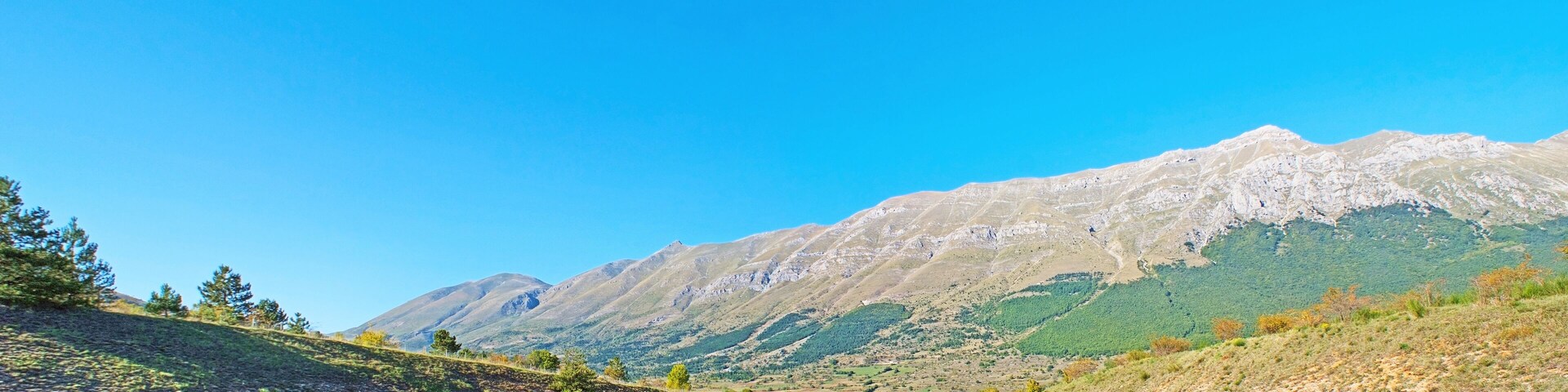 Uma estrada de montanha sinuosa com guarda-corpos, rodeada por colinas verdes e uma cadeia montanhosa imponente sob um céu azul claro.