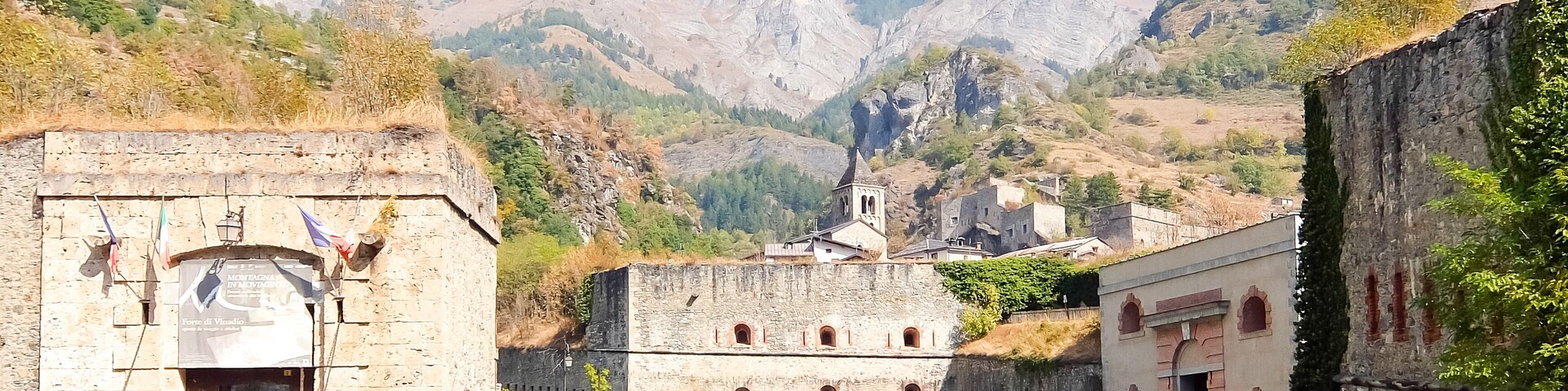 Uma fortaleza histórica de pedra com pátio, pontes em arco e montanhas ao fundo.