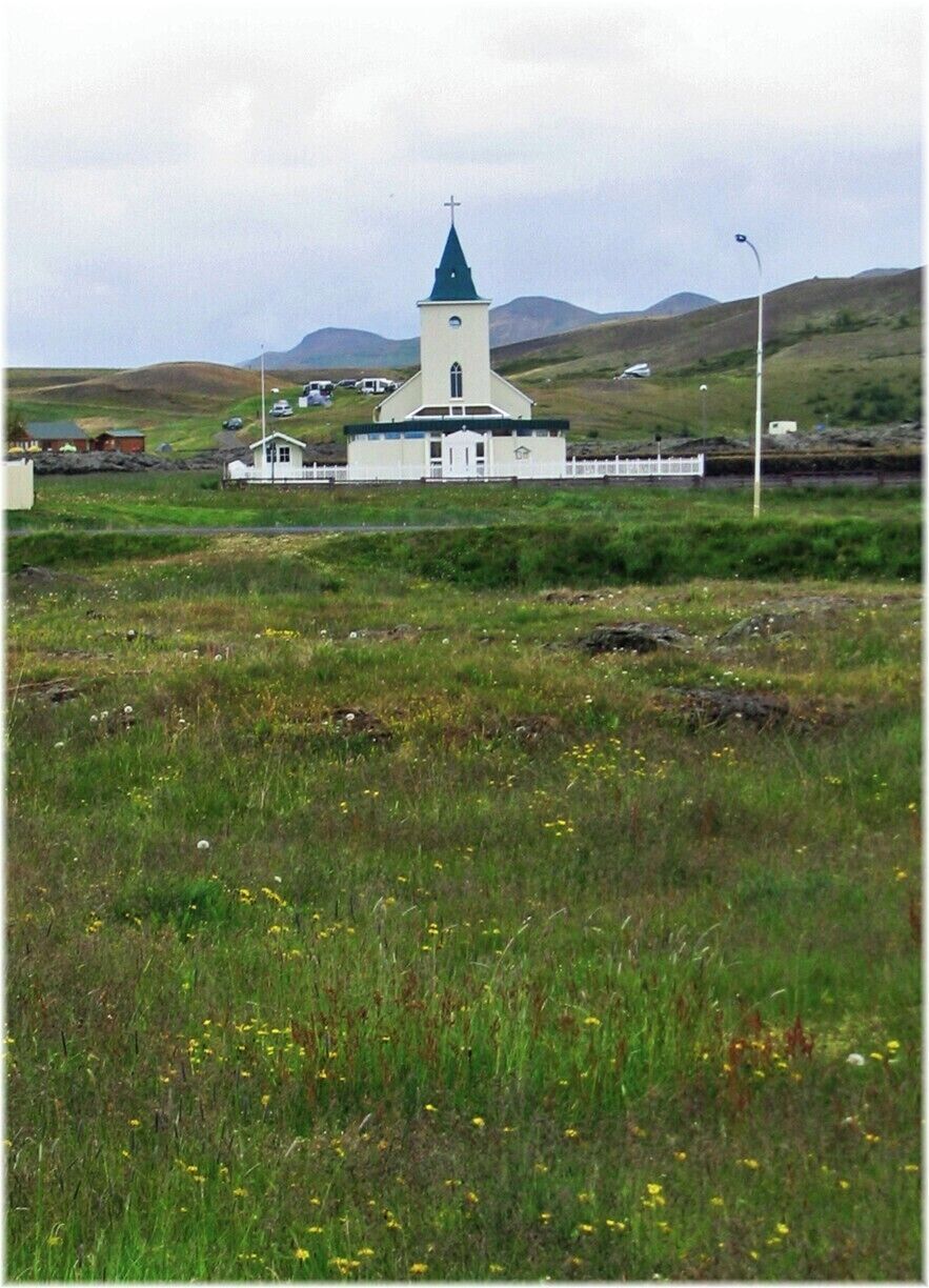 Uma igreja branca com um campanário verde e uma cruz no topo, rodeada por um campo gramado com flores amarelas e colinas ao fundo.