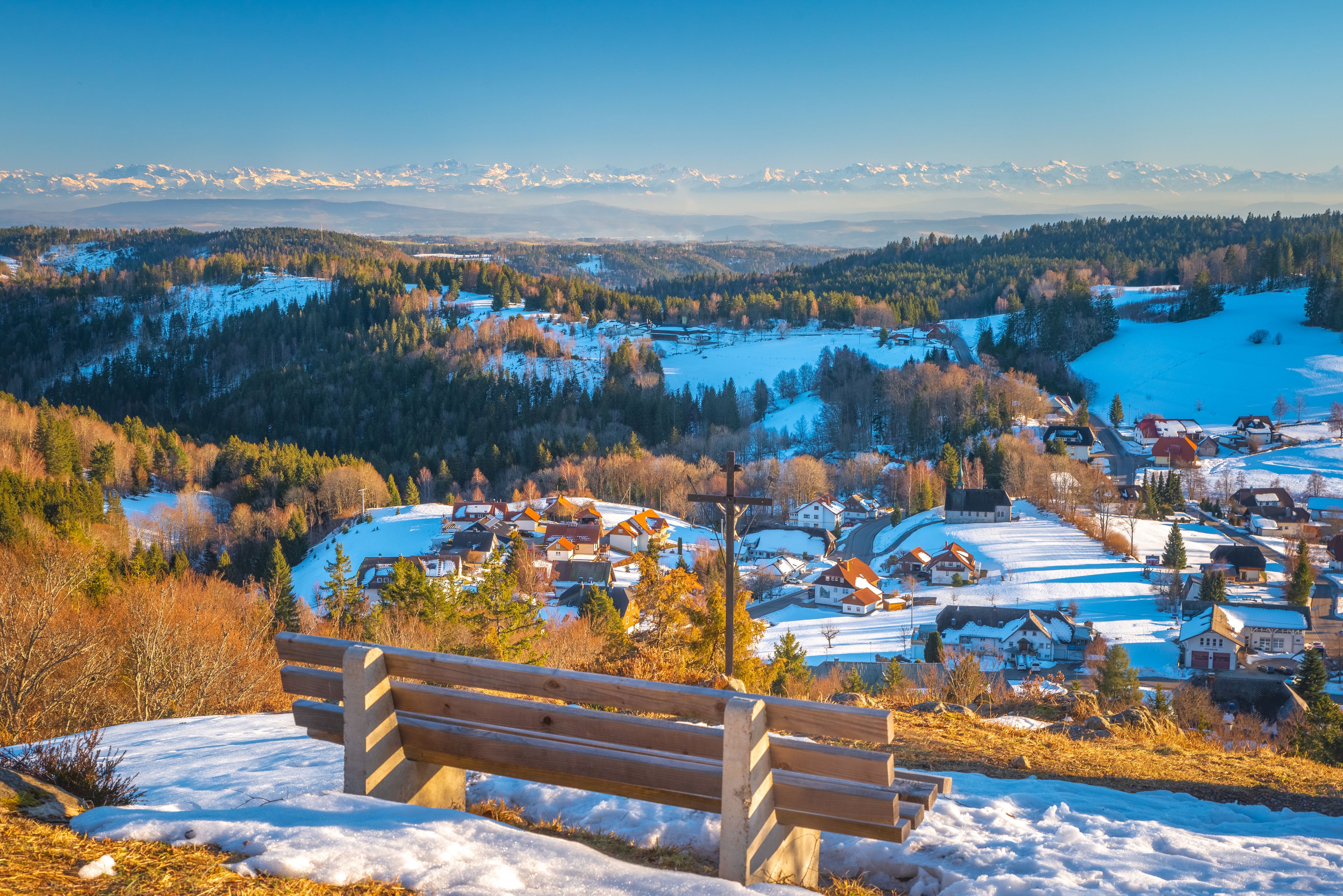 Paisagem coberta de neve com um banco de madeira, uma aldeia e montanhas ao longe.