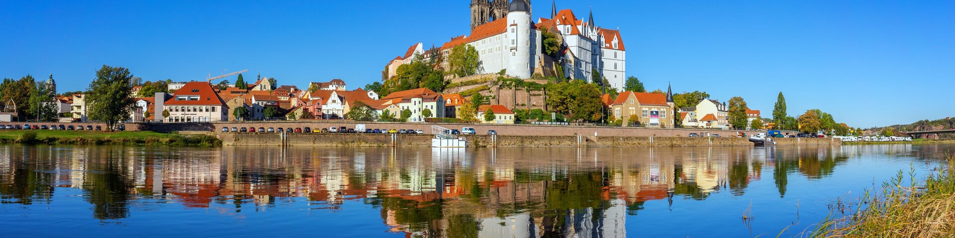 Um castelo histórico com telhados vermelhos, um rio refletindo sua imagem e um céu azul límpido.