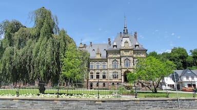 Um grande edifício histórico com arquitetura ornamentada, rodeado por um jardim bem cuidado e um muro de pedra.