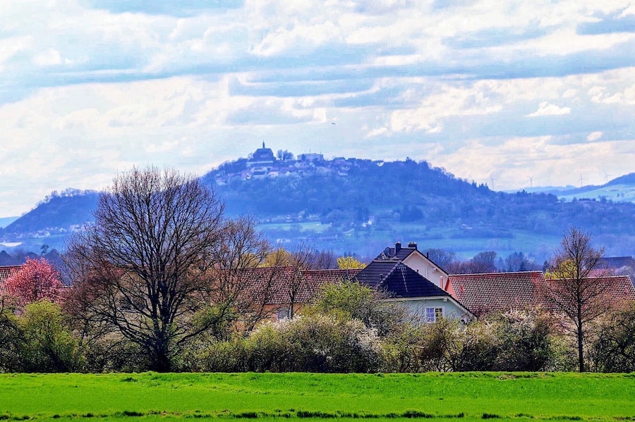 Um campo verde, uma casa com telhado vermelho e uma colina com um castelo ao fundo.