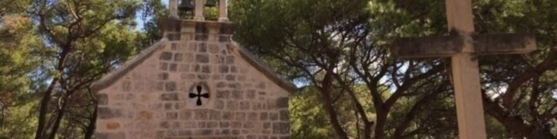 Uma pequena igreja de pedra com um campanário e uma cruz no topo, rodeada por árvores e um corpo de água.
