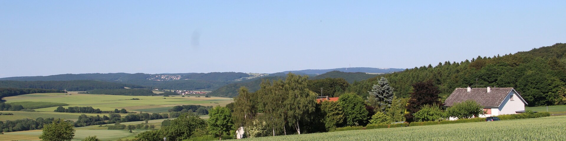 Paisagem rural com um campo de plantações, uma casa e colinas suaves ao fundo.
