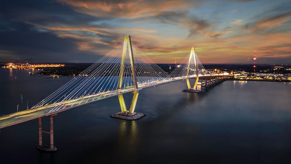 Uma ponte suspensa ao anoitecer, com cabos e torres iluminados, atravessando um corpo de água.